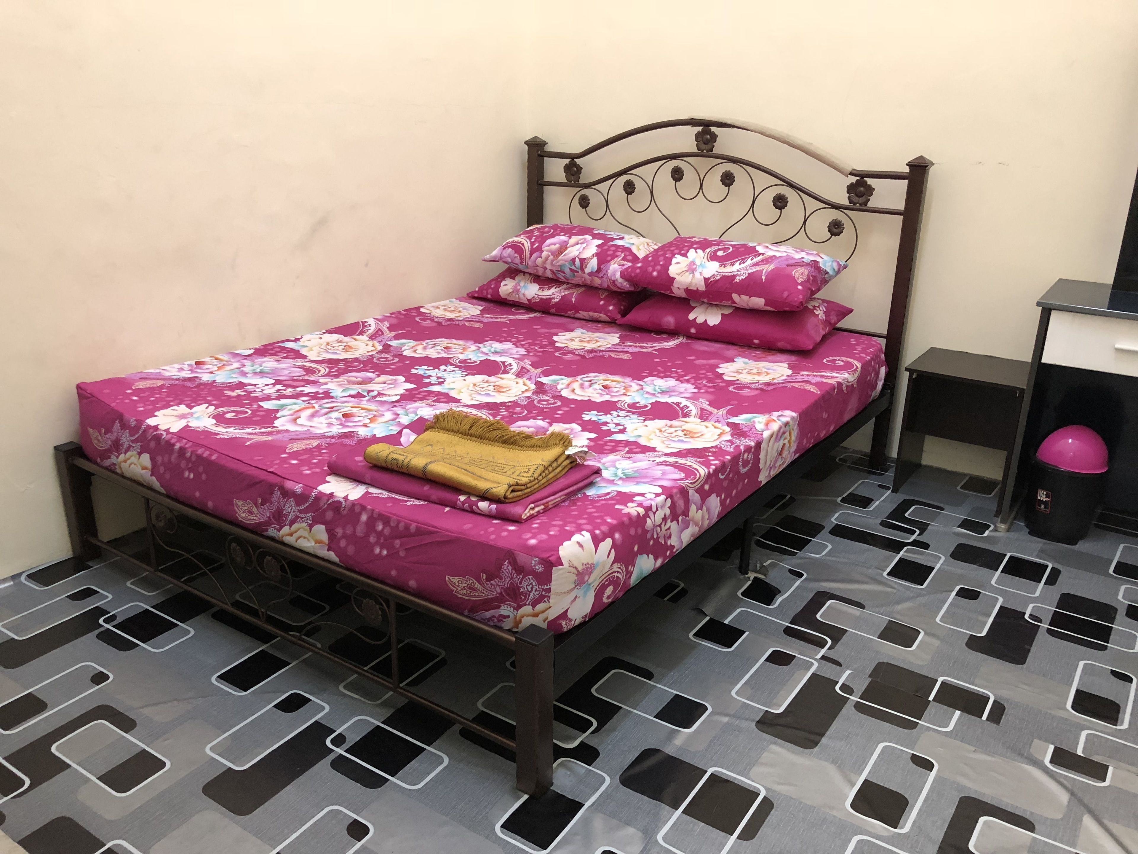 Uma cama de solteiro com colcha e almofadas florais, uma mesinha de cabeceira e uma lixeira.