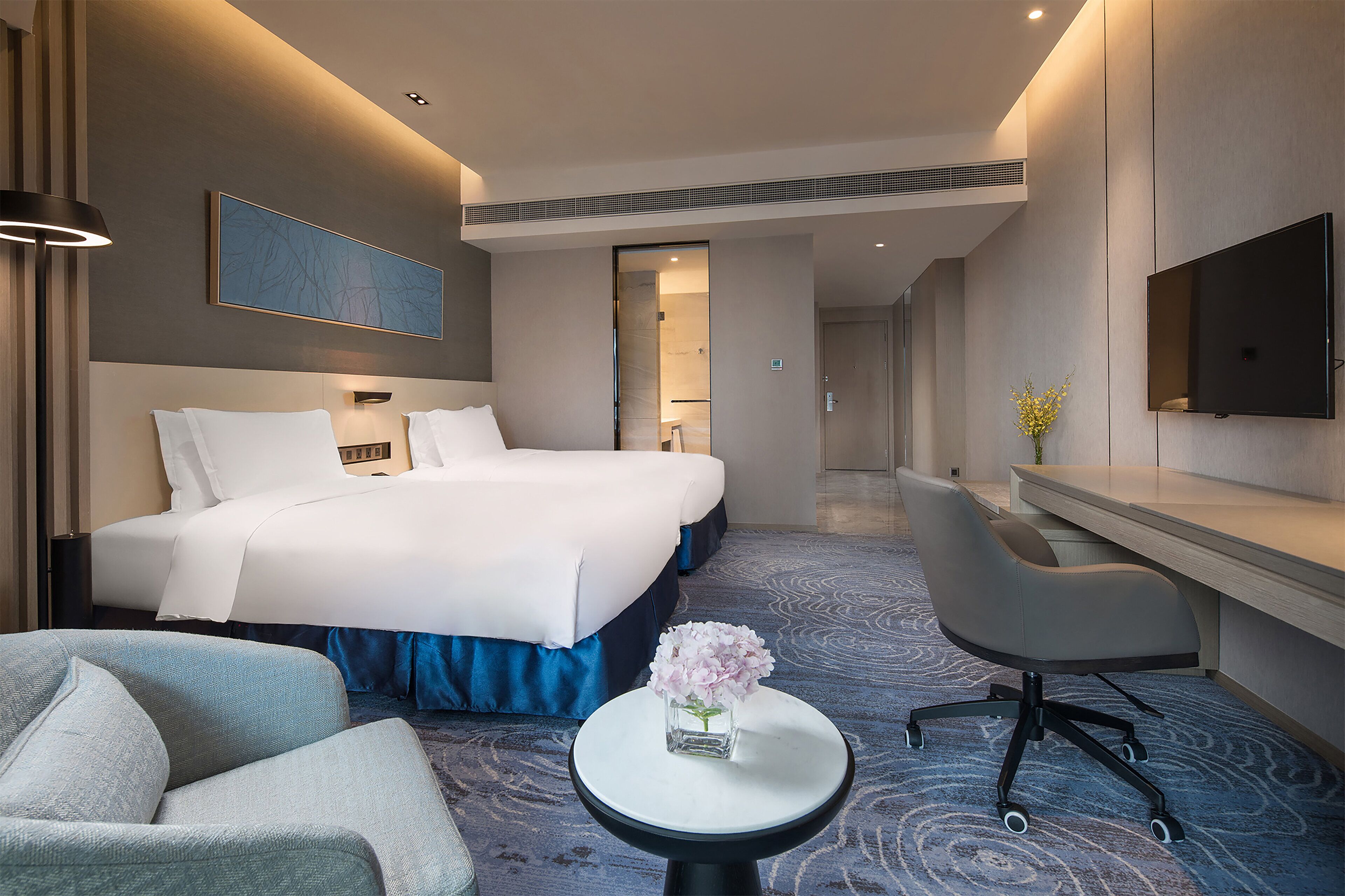 Quarto de hotel moderno com uma cama grande, uma escrivaninha, uma cadeira e uma pequena mesa com um vaso de flores.