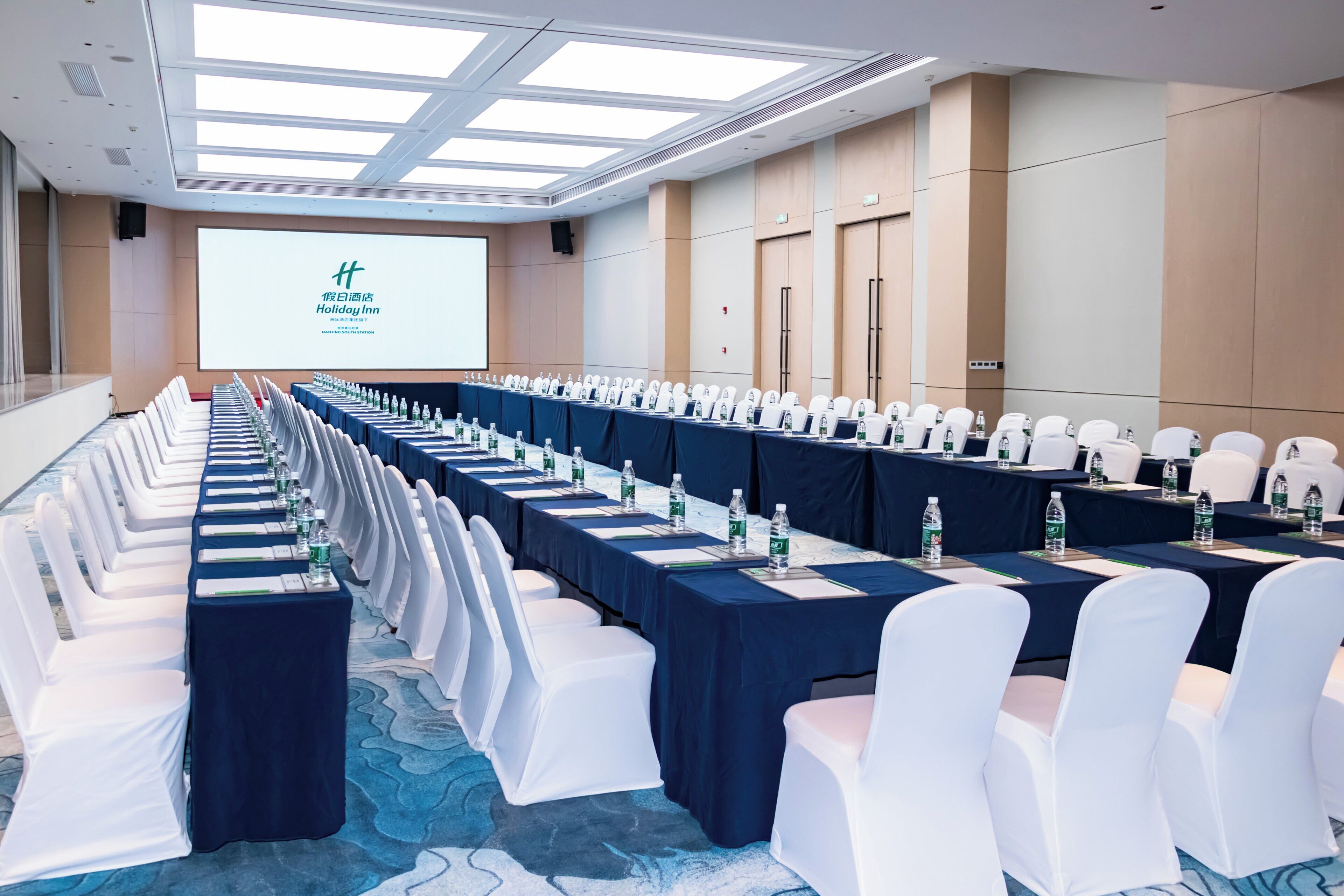 Uma sala de conferências com mesas dispostas em fileiras, cadeiras brancas e uma tela grande exibindo a marca 'Holiday Inn'.
