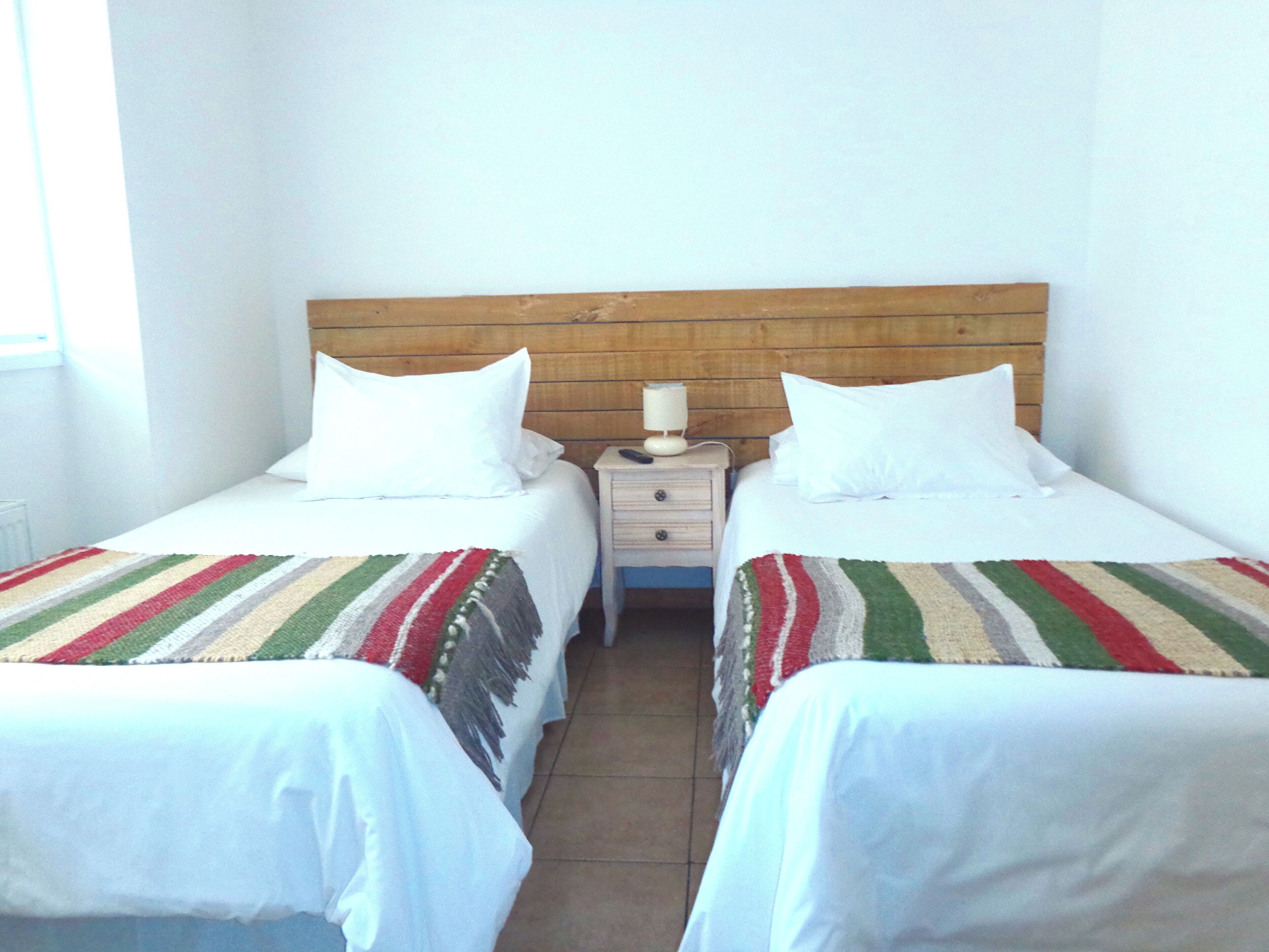 Quarto com duas camas, cada uma com roupa de cama branca e cobertores listrados. Tem uma cabeceira de madeira, uma mesa de cabeceira com uma luminária e uma janela com persianas.