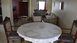 Uma área de refeições com uma mesa redonda coberta por um mantel rendado, rodeada por cadeiras de madeira com almofadas estampadas.