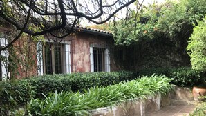 Um pátio com caminho de concreto, um pequeno edifício com janelas gradeadas e vegetação densa.