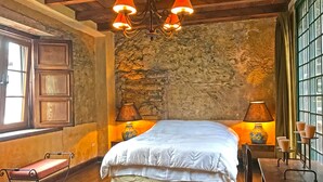 Quarto com parede de pedra, piso de madeira, cama com roupa de cama branca, duas luminárias de cabeceira, um banquinho e uma mesa com um vaso.