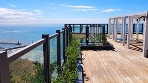 Um deck de madeira com guarda-corpos de vidro, vista para o mar, um pequeno espelho d'água e área de estar com vista para o oceano.