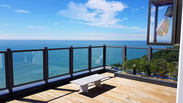 Uma varanda com piso de madeira, um banco de pedra e guarda-corpo de vidro com vista para o mar.