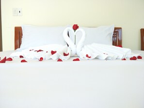 Uma cama com lençóis brancos, arranjo de toalha em formato de coração e pétalas de rosa espalhadas.