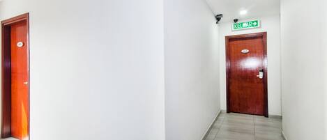 Um corredor com uma porta de madeira à esquerda e uma placa de saída verde sobre outra porta à direita.
