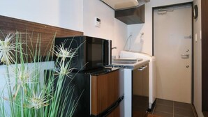 Uma cozinha compacta com micro-ondas, fogão e pia. Há uma planta em vaso com folhas verdes longas. A cozinha possui armários de madeira e bancada branca.