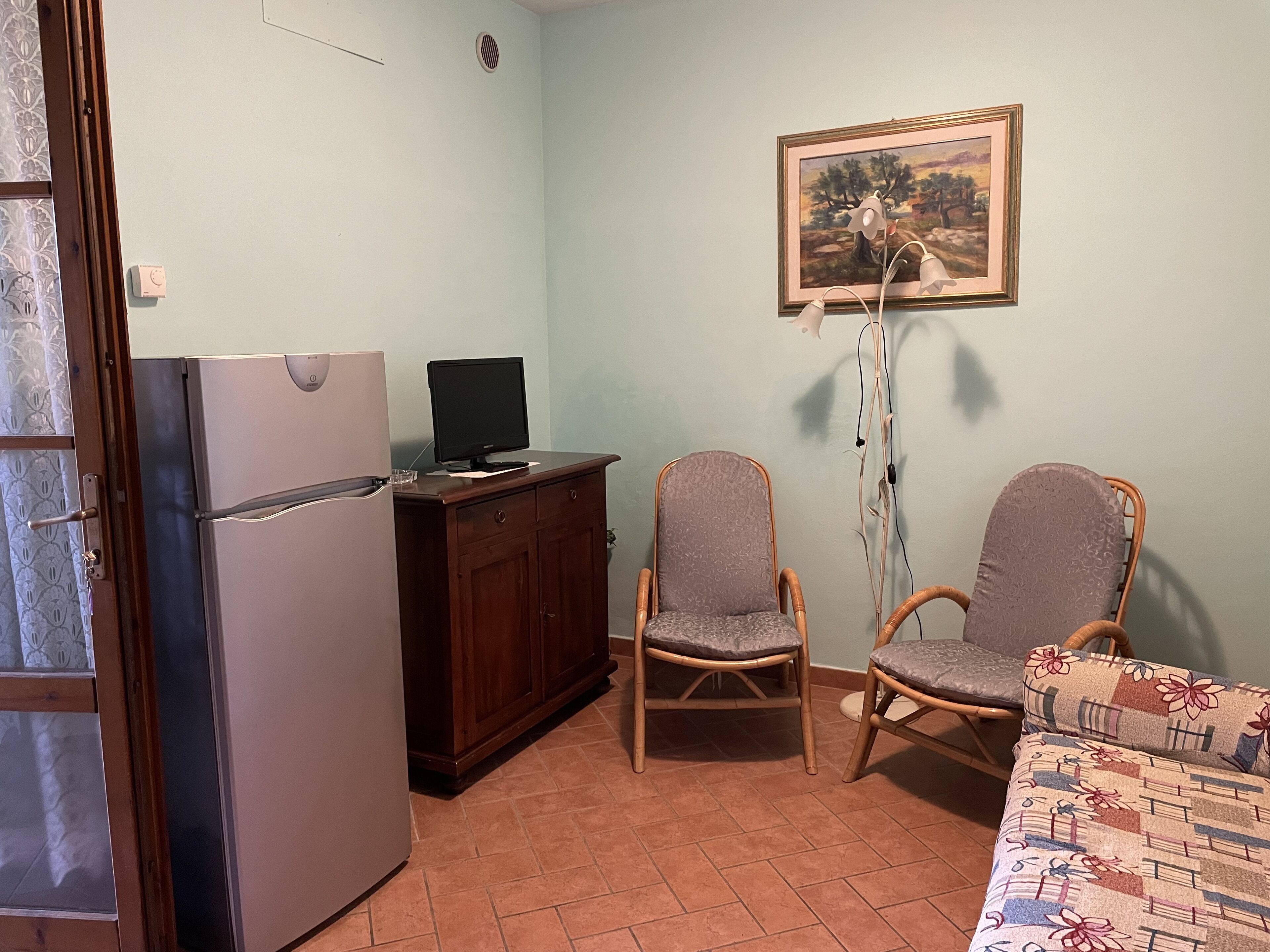 Quarto compacto com geladeira, um armário de madeira, duas cadeiras de vime e um quadro na parede.