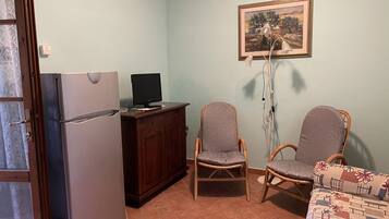 Quarto compacto com geladeira, um armário de madeira, duas cadeiras de vime e um quadro na parede.
