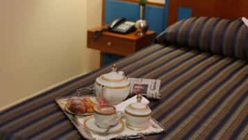Quarto de hotel com uma cama, mesa de cabeceira com telefone, vaso com flores e bandeja com itens de café da manhã.