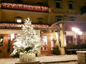 Entrada de hotel com uma árvore coberta de neve em primeiro plano, fachada com iluminação acolhedora e uma placa com a inscrição 