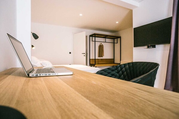 Quarto de hotel moderno com escrivaninha de madeira, um laptop, uma cadeira preta, cama com roupa de cama branca, cabides e uma TV montada.