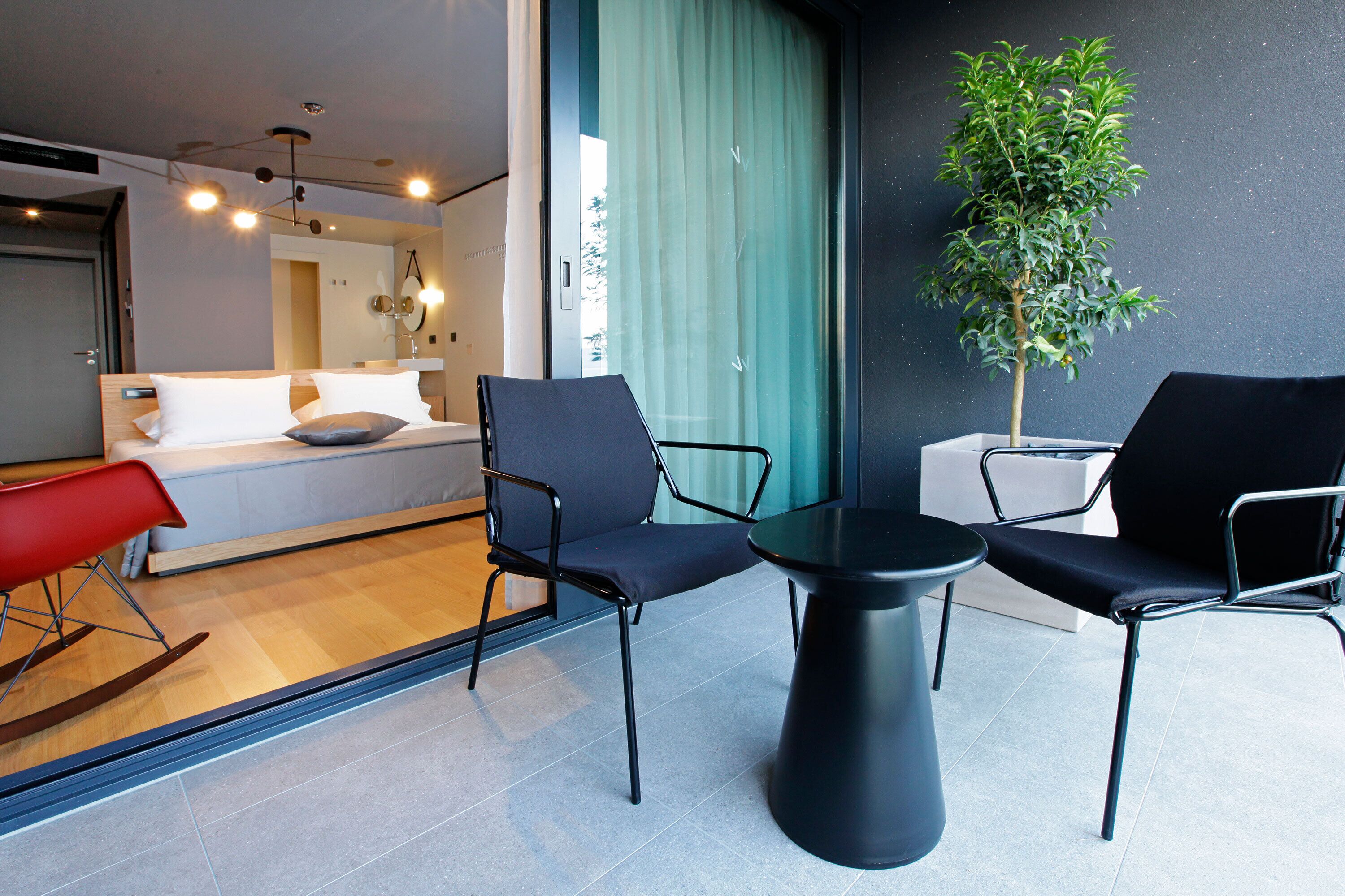 Quarto de hotel moderno com uma cama, uma cadeira vermelha, duas cadeiras pretas, uma mesinha e um vaso com planta.