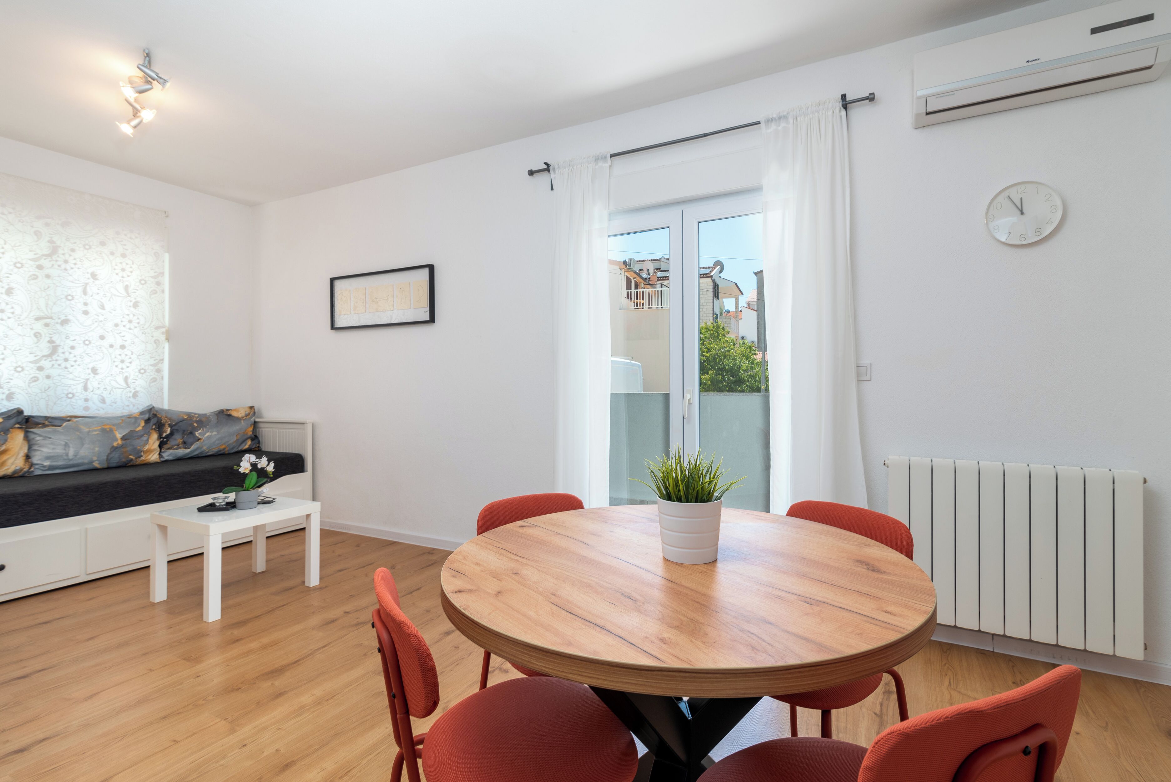 Uma sala de estar moderna com uma mesa de madeira redonda, cadeiras laranja, um sofá branco e um vaso com planta.