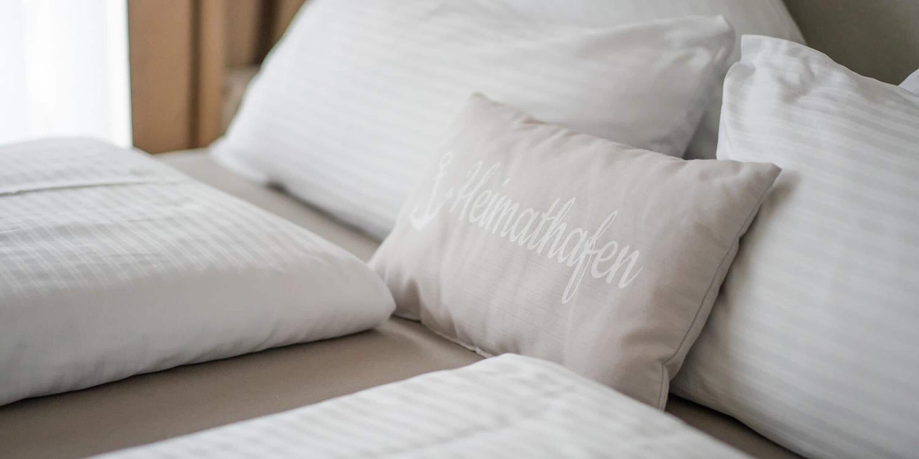 Uma cama com roupa de cama e travesseiros brancos, um dos travesseiros tem texto.