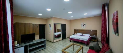Quarto de hotel com cama, televisão, sofá, mesinha e espelho.