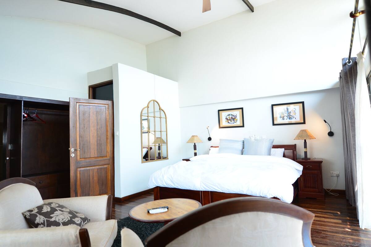 Quarto de hotel com cama, guarda-roupa de madeira, cadeira, mesa redonda pequena e abajur.