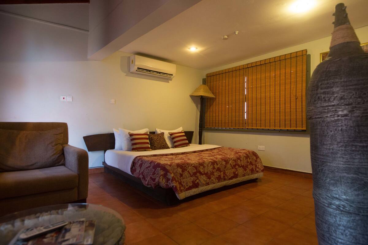 Quarto com cama, sofá, ar condicionado, janela com persianas e luminária.
