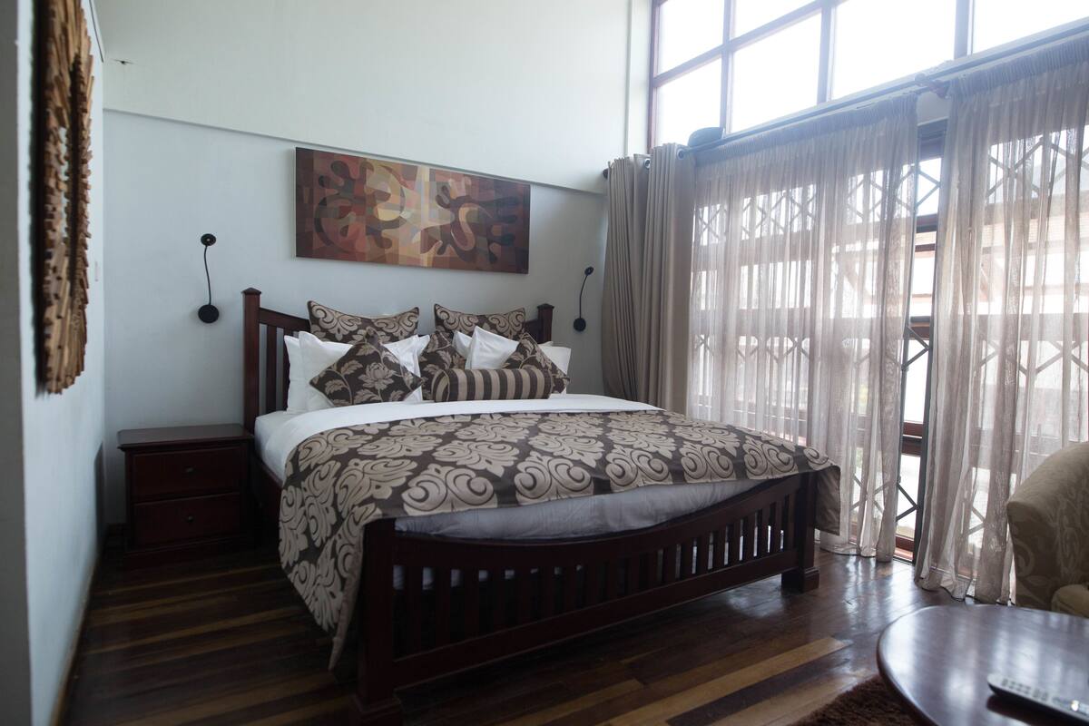Quarto com cama de madeira, mesa de cabeceira, luminária na parede e uma janela grande com cortinas translúcidas.