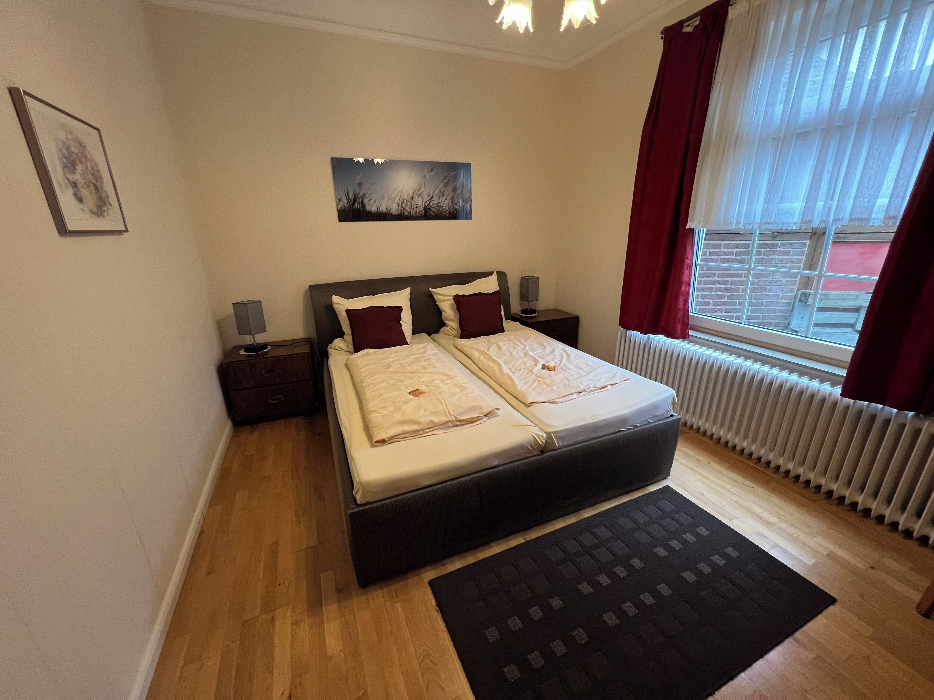 Quarto com cama grande, duas mesas de cabeceira, um criado-mudo, um radiador e uma janela com cortinas.