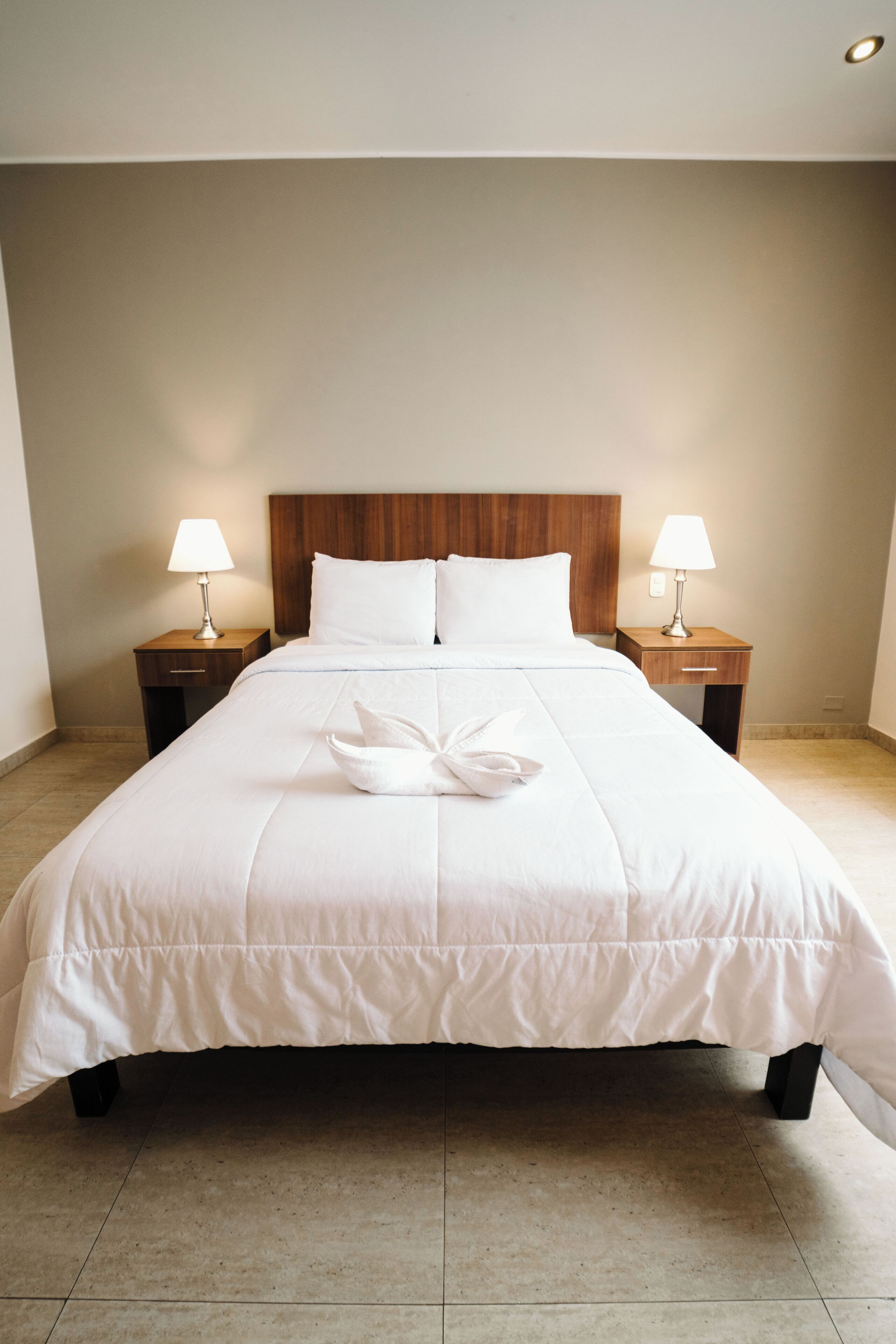 Cama bem arrumada com roupa de cama branca e almofadas decorativas. Há duas mesas de cabeceira com abajures, e o quarto tem um design simples e limpo.