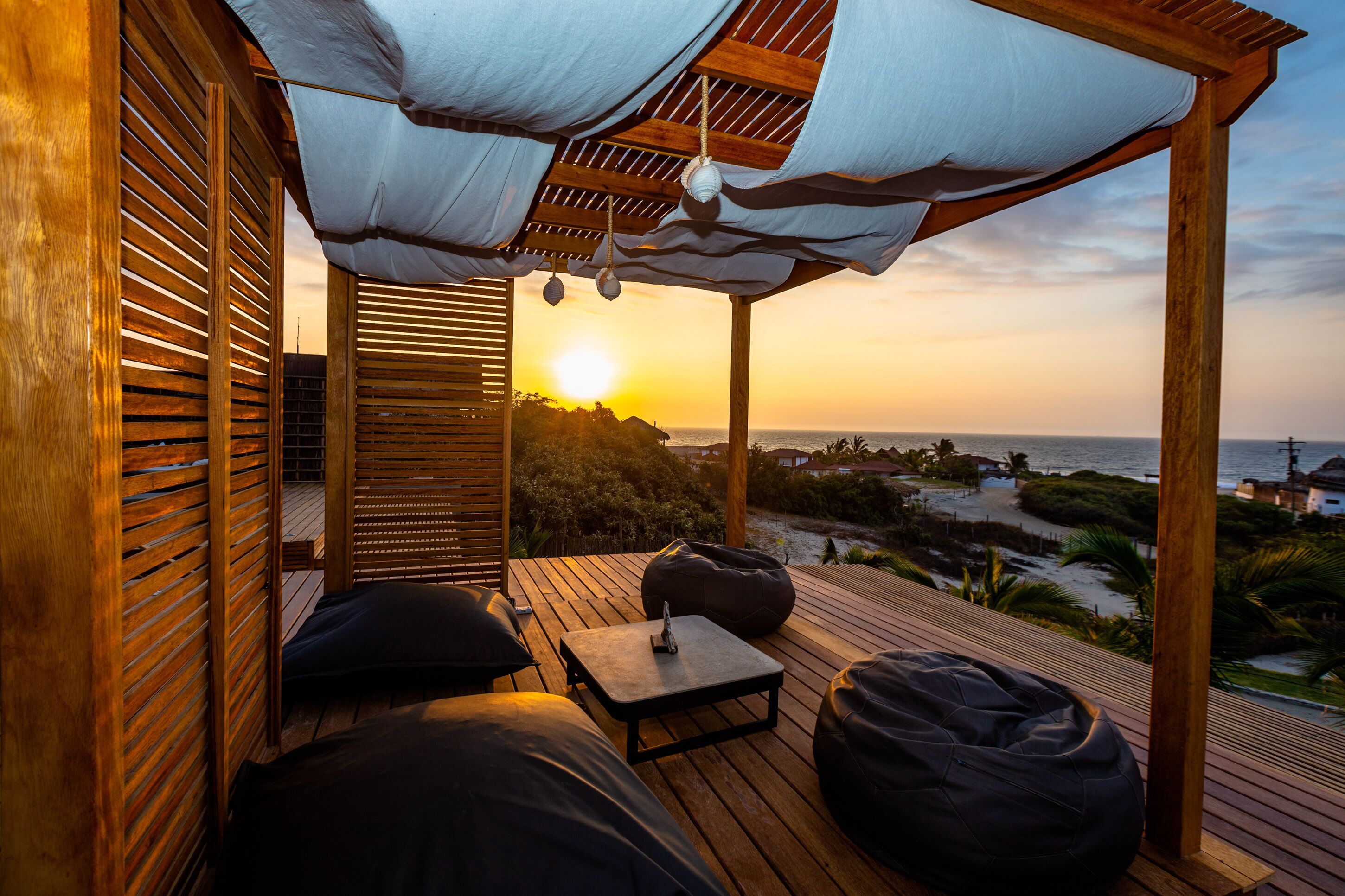 Um deck de madeira com uma mesa e poltronas de bean bag, com vista para uma praia ao pôr do sol.