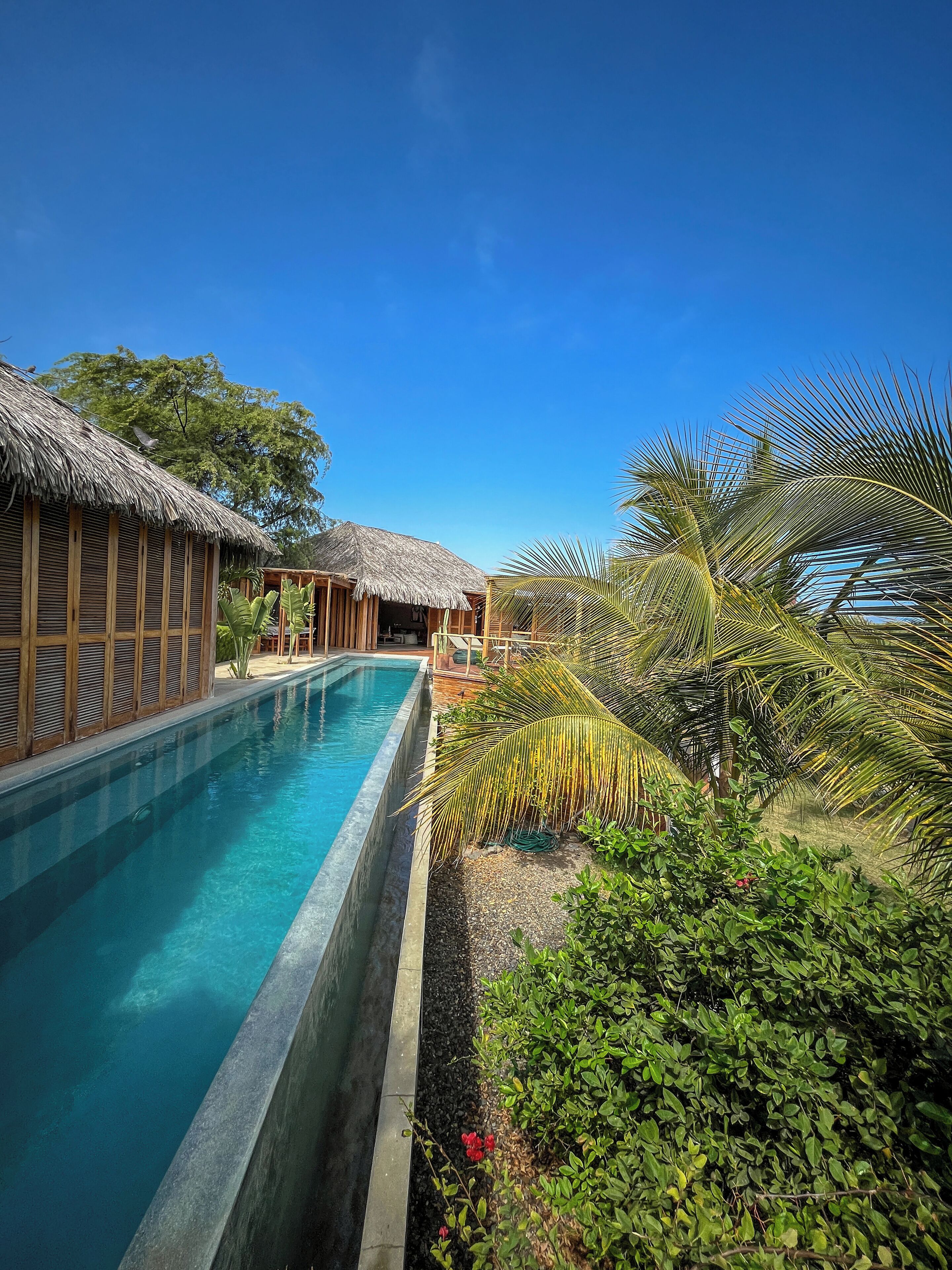Uma área de piscina com piscina azul cristalina, estruturas de madeira com telhados de palha e vegetação tropical.