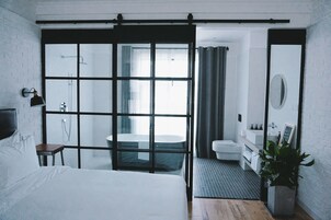 Quarto moderno com uma cama grande, uma mesa de cabeceira, uma luminária e uma janela com vista para um banheiro, além de uma planta em vaso.