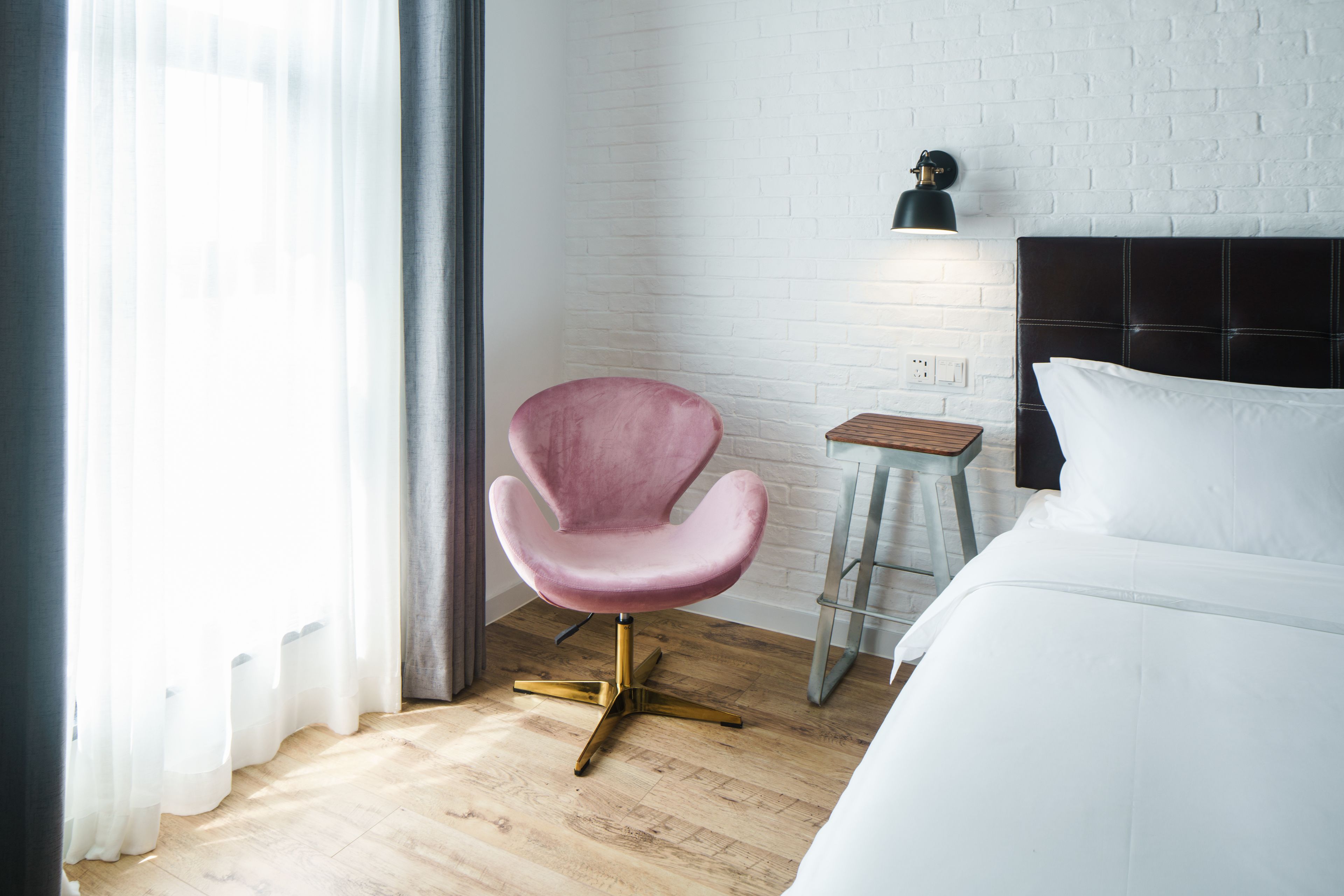 Quarto de hotel com uma cama, uma cadeira rosa, uma mesinha, uma luminária e uma parede de tijolos brancos.