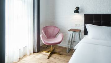 Quarto de hotel com uma cama, uma cadeira rosa, uma mesinha, uma luminária e uma parede de tijolos brancos.