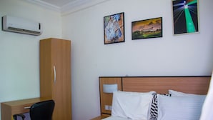 Quarto de hotel com cama, escrivaninha, cadeira e ar condicionado. Três quadros emoldurados estão pendurados na parede.