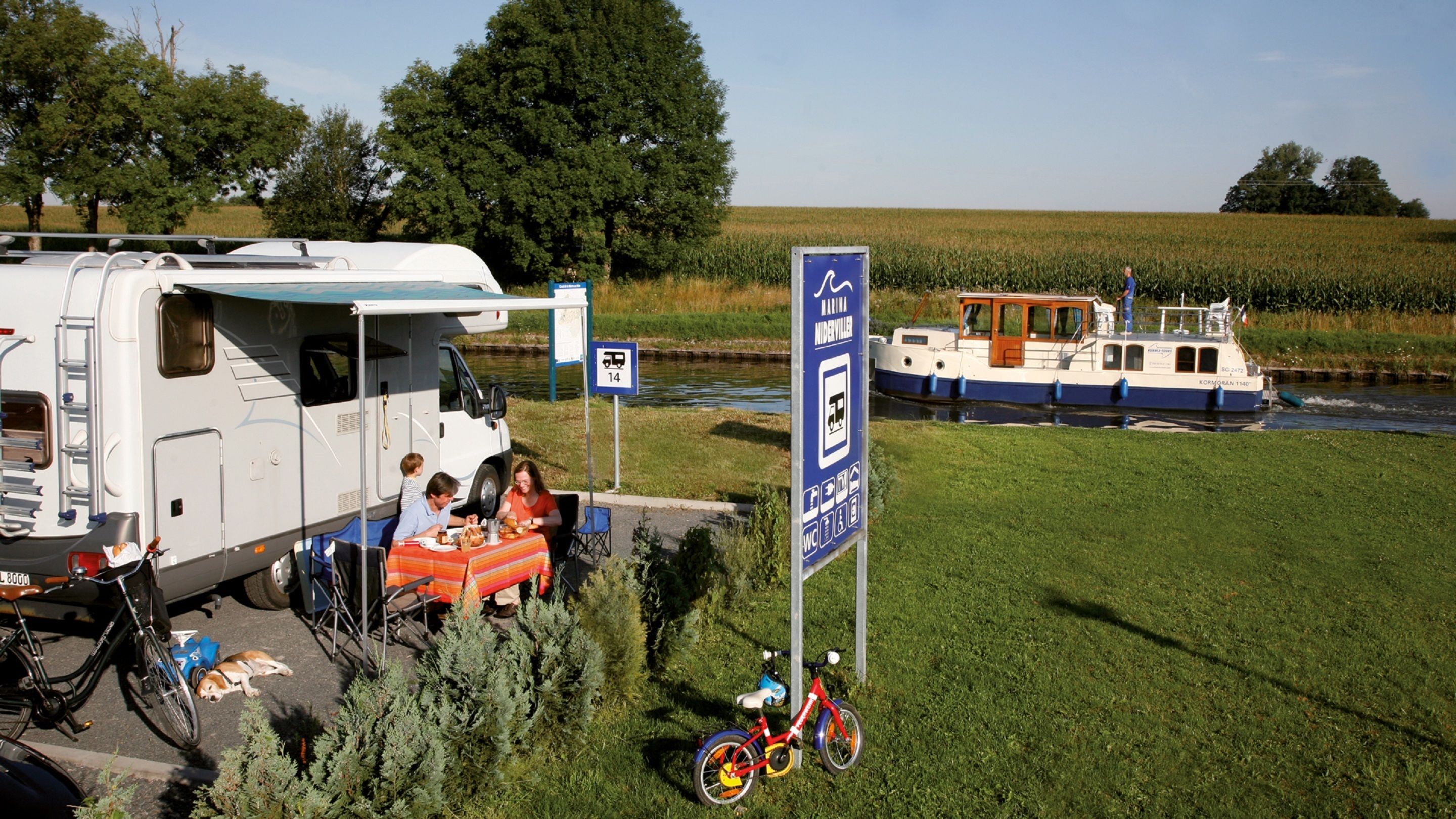 Um camping com uma RV branca, uma mesa de jantar com pessoas, uma bicicleta e um barco na água.