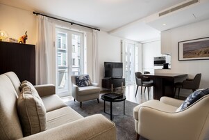 Uma sala de estar moderna com sofá, poltrona e uma pequena mesa de centro. Há uma televisão e uma área de jantar com cadeiras.