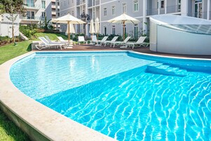 Piscina com espreguiçadeiras e guarda-sóis, cercada por um gramado bem cuidado e um edifício com varandas.