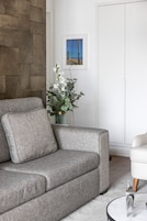 Uma sala de estar moderna com um sofá cinzento, uma mesa pequena com um objeto decorativo, um vaso com flores e um quadro na parede.