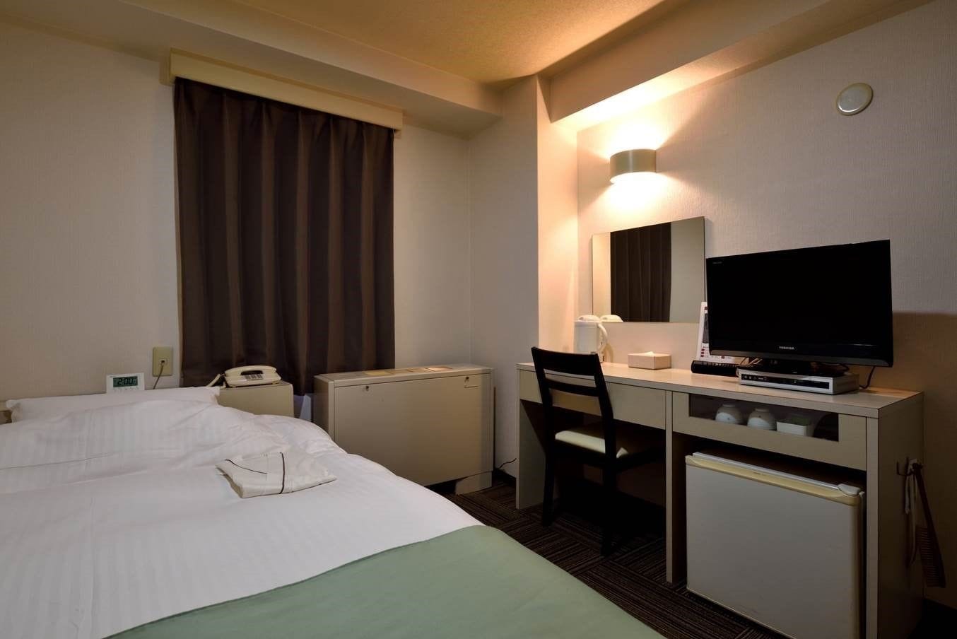 Quarto de hotel com uma cama, uma escrivaninha com televisão, uma cadeira, abajur e um pequeno frigorífico.