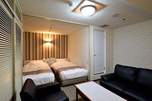 Quarto de hotel com duas camas, um sofá de couro preto, uma mesa pequena e uma porta.