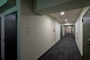 Corredor longo e estreito de um hotel, com portas em ambos os lados, chão coberto de carpete e teto com iluminação embutida.