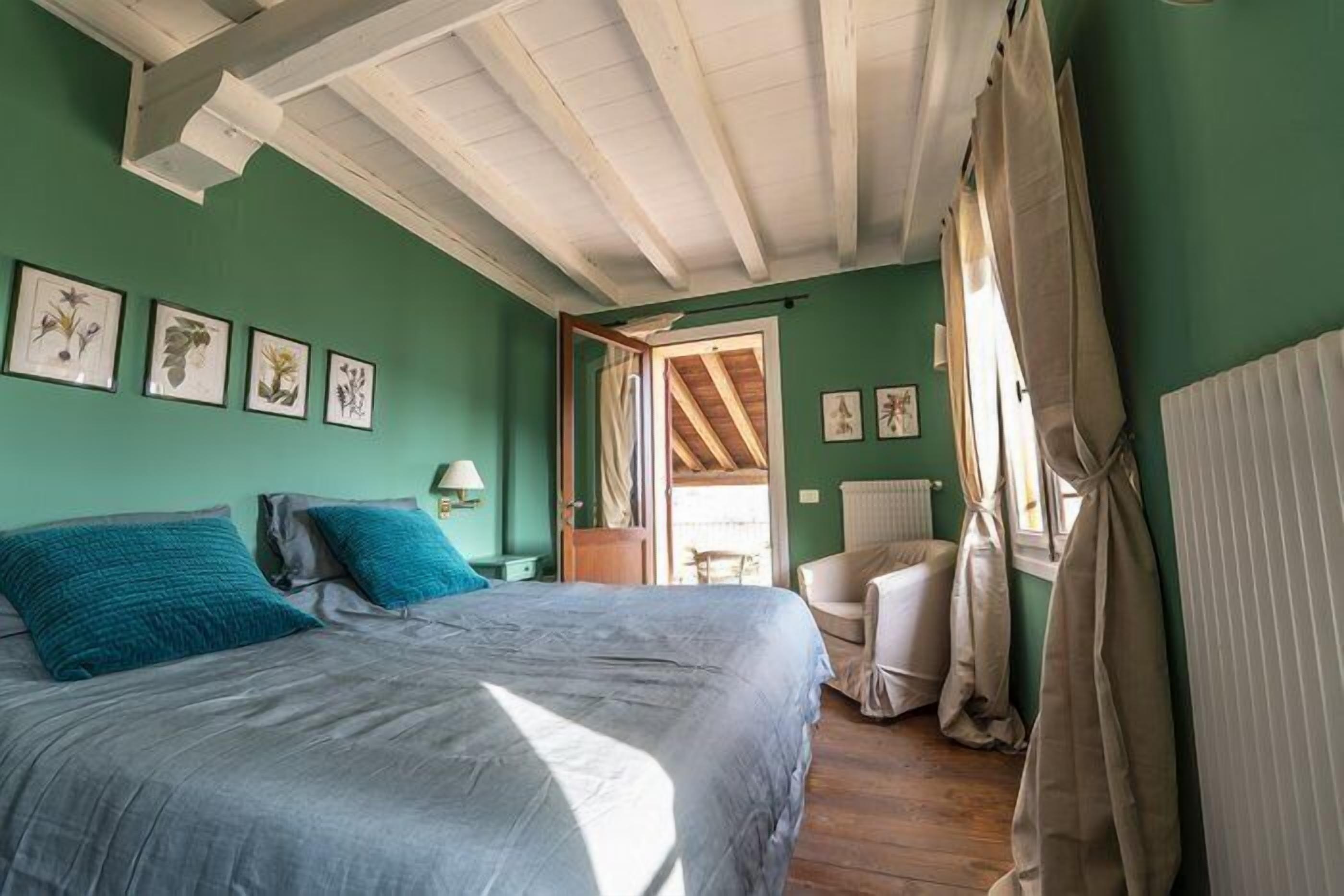 Quarto com cama, cadeira, janela com cortinas e gravuras de plantas emolduradas na parede.