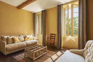 Um quarto com sofá bege, mesa de centro de madeira, cadeira de madeira e uma grande janela com cortinas.