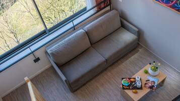 Uma sala de estar moderna com um sofá, uma pequena mesa de madeira com uma planta e uma janela com vista para árvores.