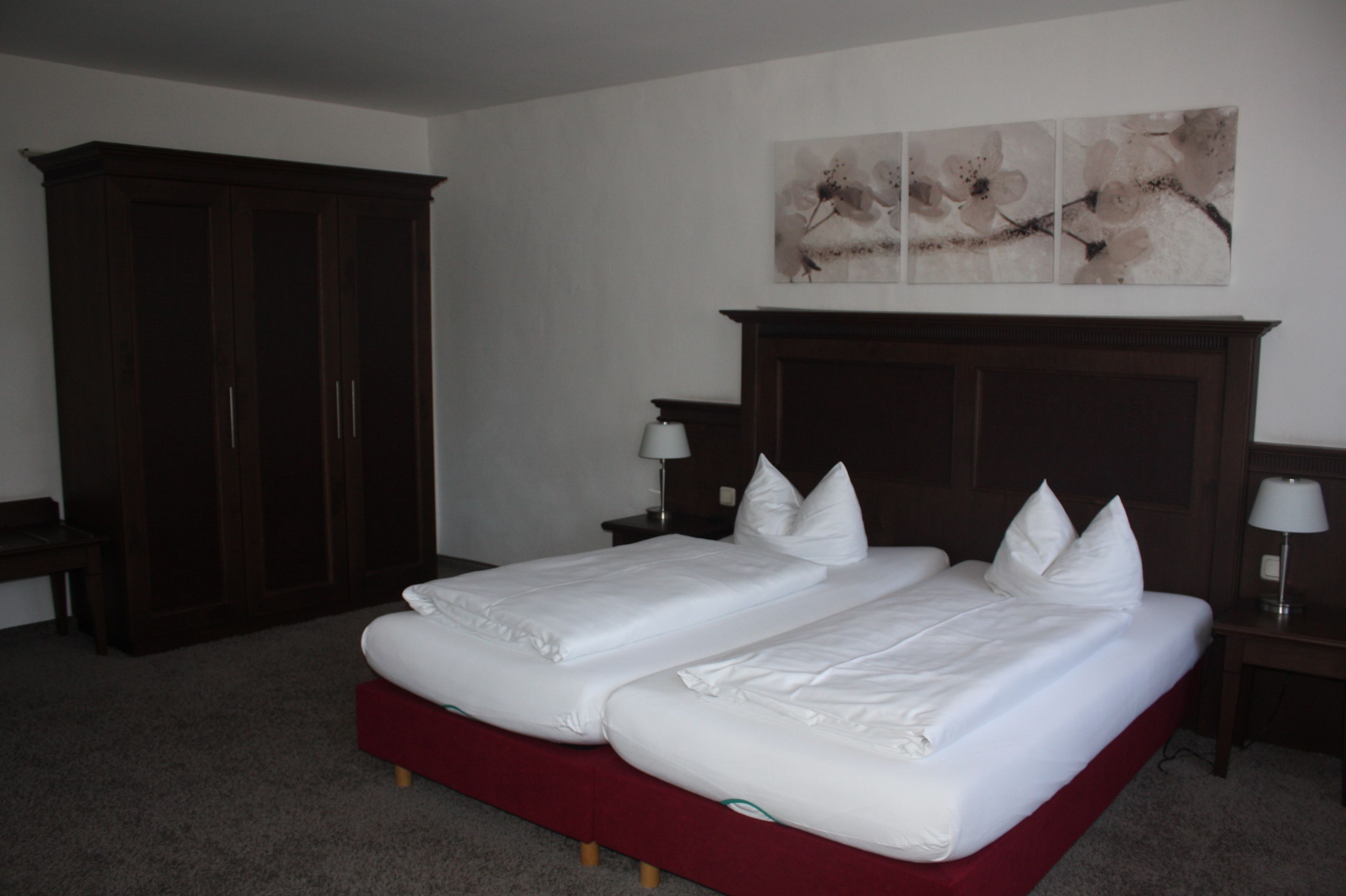 Cama de casal com lençóis brancos e base vermelha, duas mesas de cabeceira com abajures, guarda-roupa de madeira escura e uma obra de arte floral pendurada na parede.