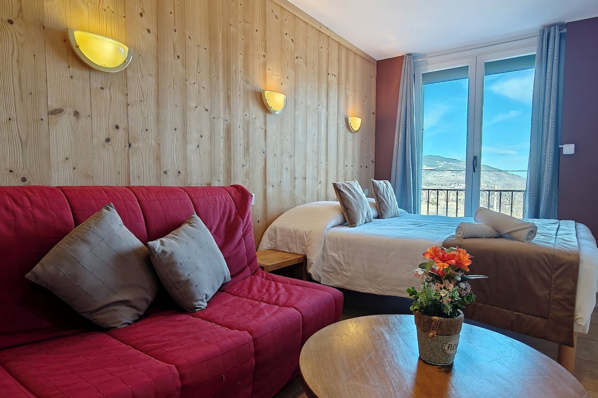 Quarto de hotel com parede de madeira, duas camas, um sofá vermelho, uma mesa de madeira redonda e uma planta em vaso.