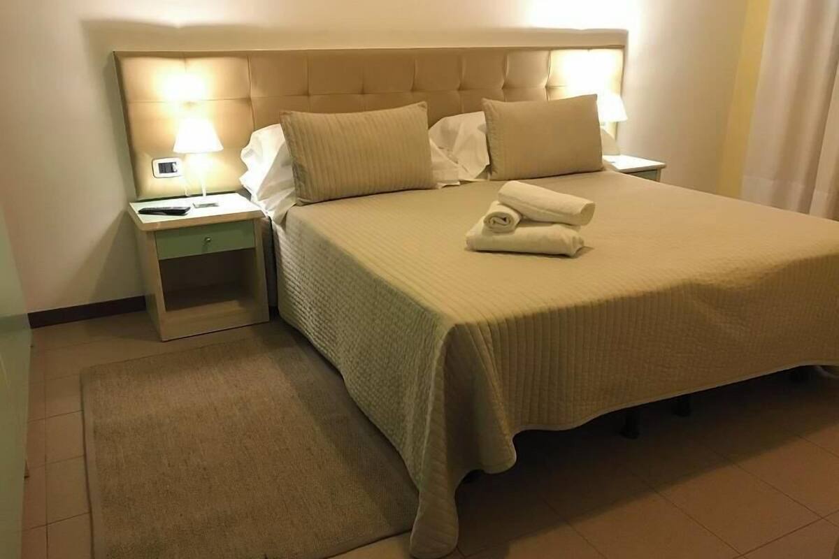 Cama bem arrumada com colcha bege e travesseiros, mesa de cabeceira com abajur e um criado-mudo.