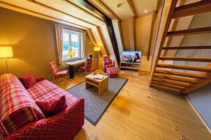 Uma sala de estar acolhedora com um sofá de retalhos vermelhos, uma mesa de centro de madeira e uma televisão. Há uma janela com cortinas, uma escrivaninha com uma luminária e uma escada de madeira.