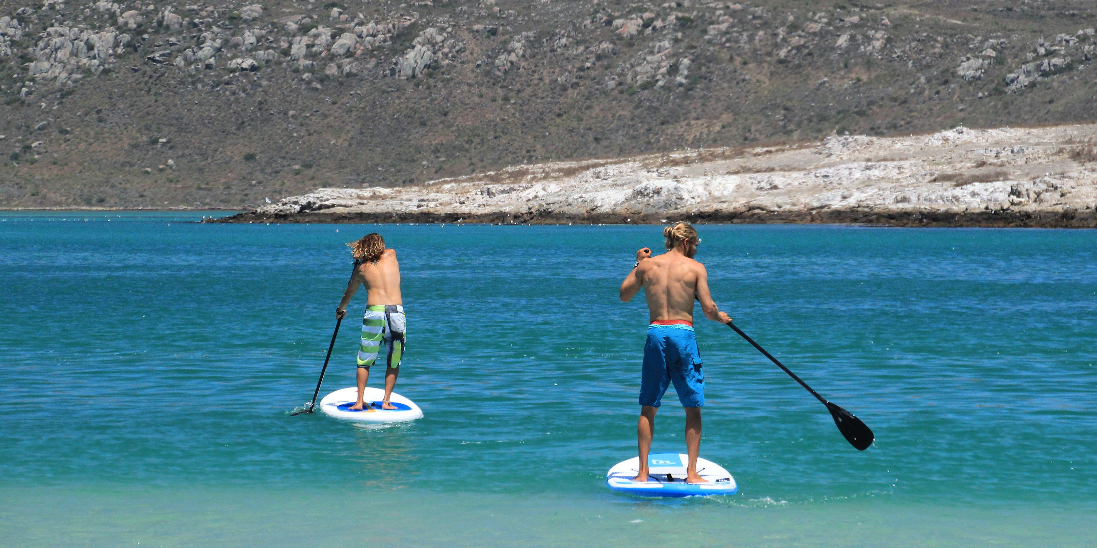 Duas pessoas praticando paddleboarding em um mar calmo, com colinas rochosas ao fundo.