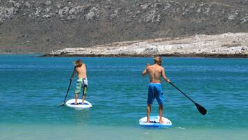 Duas pessoas praticando paddleboarding em um mar calmo, com colinas rochosas ao fundo.