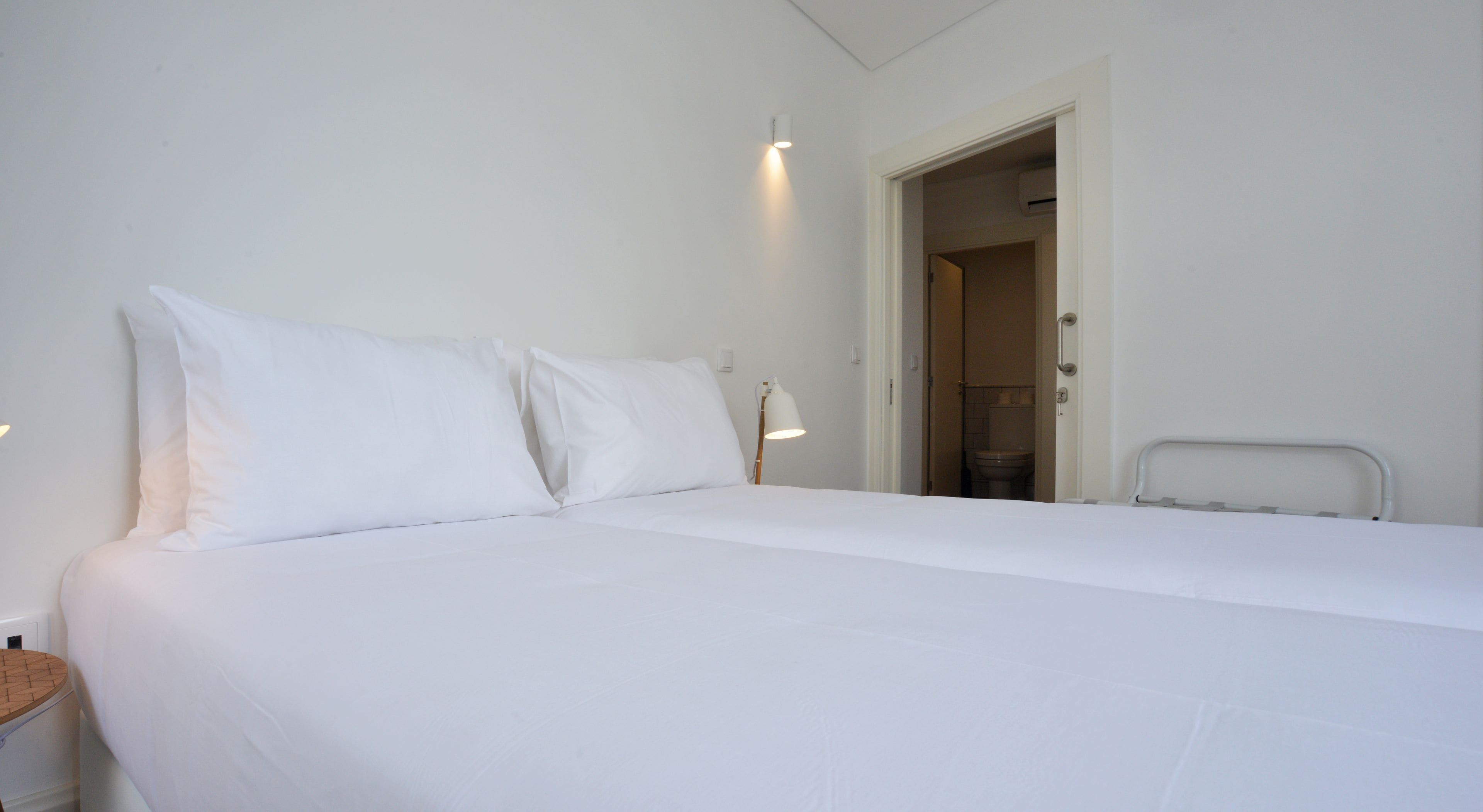 Cama bem arrumada com lençóis brancos num quarto com paredes brancas e uma porta que dá para o banheiro.
