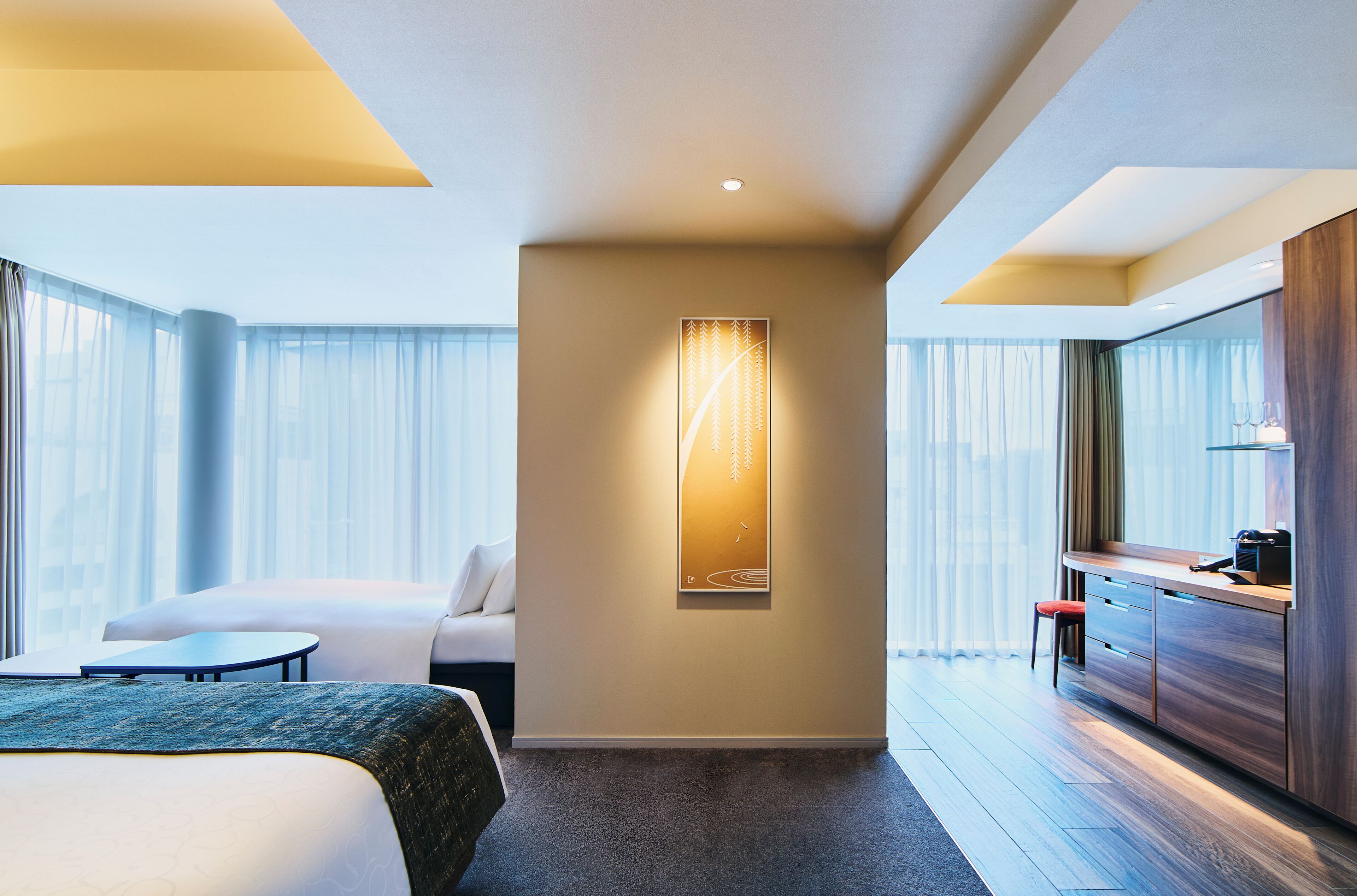 Quarto de hotel com cama, escrivaninha, poltrona, sofá e uma janela grande com cortinas.